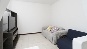 Uma sala de estar pequena e moderna com uma TV de tela plana em um suporte de madeira escura, um sofá cinza com almofadas estampadas e uma poltrona azul.