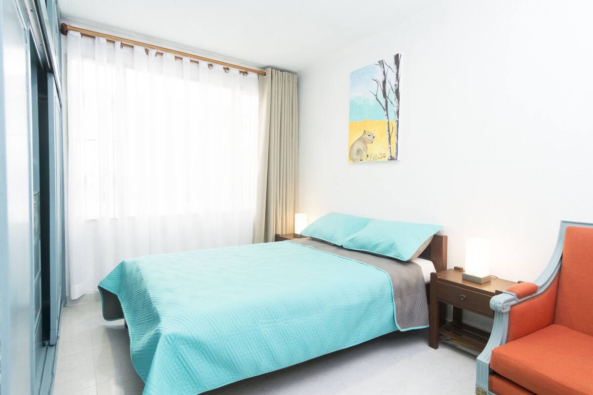 Quarto com cama, mesa de cabeceira, cadeira e um quadro na parede.