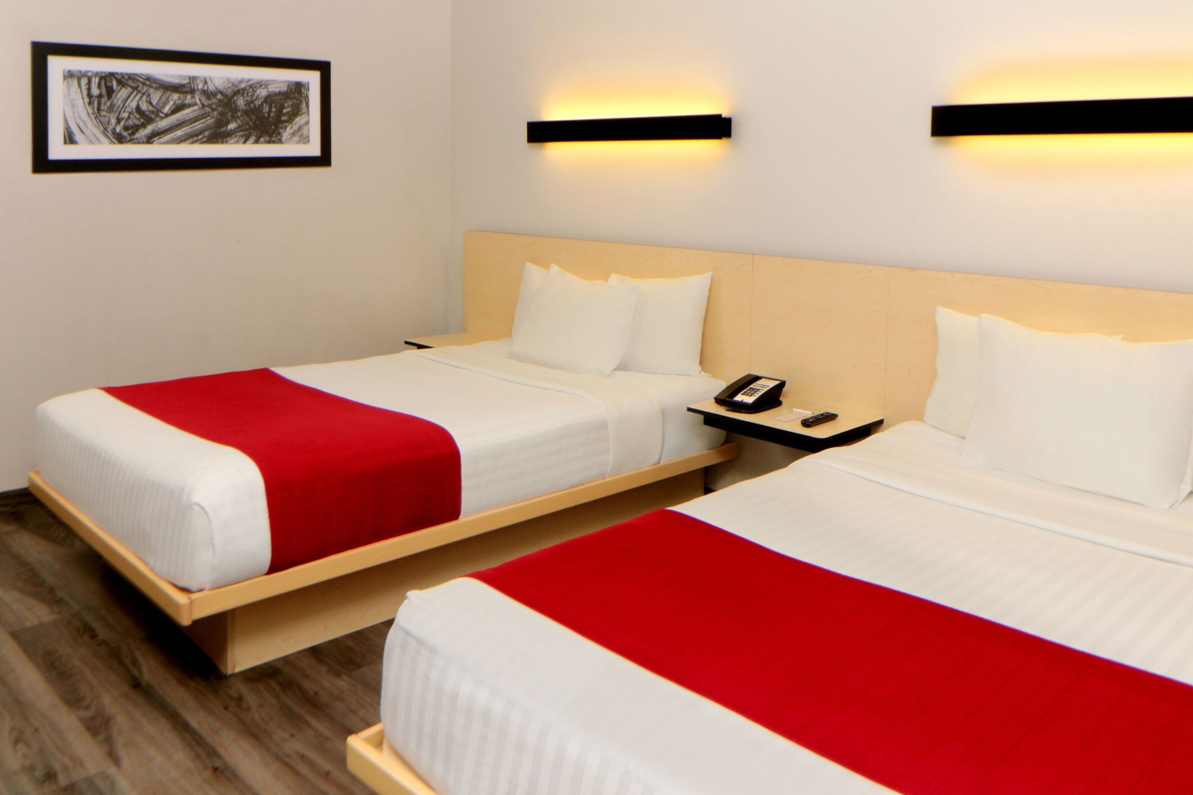Quarto de hotel com duas camas, cada uma com colcha vermelha e branca, cabeceira de madeira e mesa de cabeceira com telefone e controle remoto.