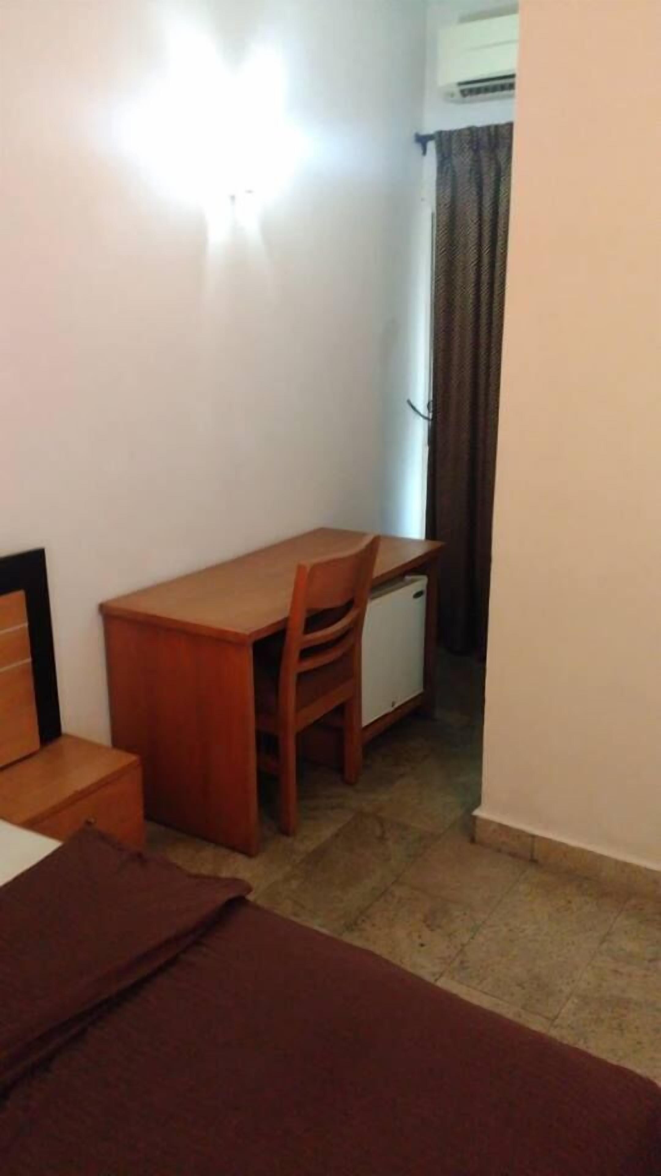 Quarto pequeno e compacto com uma escrivaninha de madeira, uma cadeira, uma cama e um armário pequeno e branco.