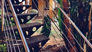 Uma escadaria em espiral com corrimãos de metal e área de desembarque com ladrilhos de pedra, cercada por plantas tropicais e um edifício com telhado ladrilhado.