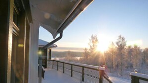 Varanda coberta de neve com guarda-corpo, com vista para uma floresta e um corpo d'água ao nascer do sol.