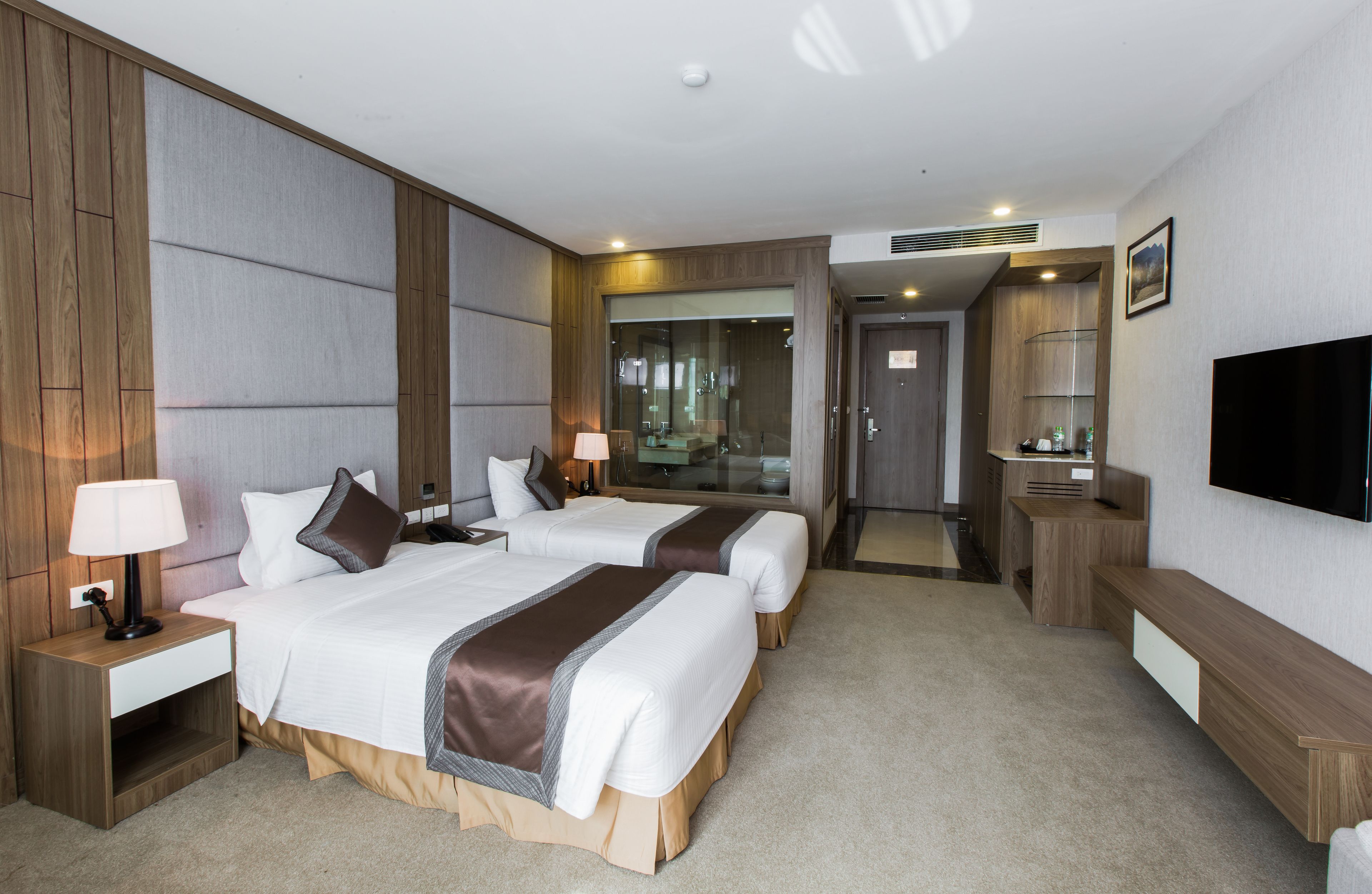 Quarto de hotel com duas camas, uma televisão, uma mesa de trabalho e um banheiro visível através de uma porta de vidro.
