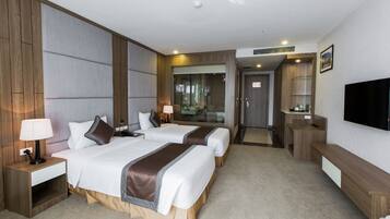 Quarto de hotel com duas camas, uma televisão, uma mesa de trabalho e um banheiro visível através de uma porta de vidro.