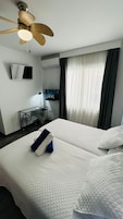 Quarto de hotel com duas camas, um ventilador de teto, televisão de tela plana, ar condicionado, uma mesa com cadeira e uma janela com cortinas.