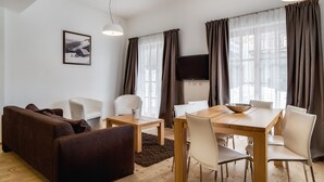 Uma sala de estar moderna com um sofá marrom, uma poltrona branca, uma mesa de jantar de madeira com cadeiras e uma TV de tela plana na parede.