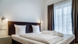 Cama bem arrumada com lençóis brancos, cabeceira escura e duas luminárias de leitura fixadas na parede.