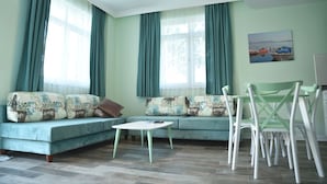 Uma sala de estar com sofá, uma mesinha e uma cadeira. Há cortinas nas janelas e um quadro na parede.