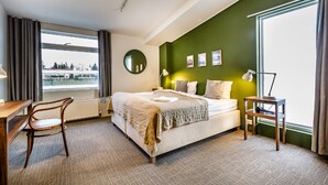 Quarto de hotel com cama, escrivaninha, cadeira, janela com cortinas, espelho redondo e parede verde decorativa.
