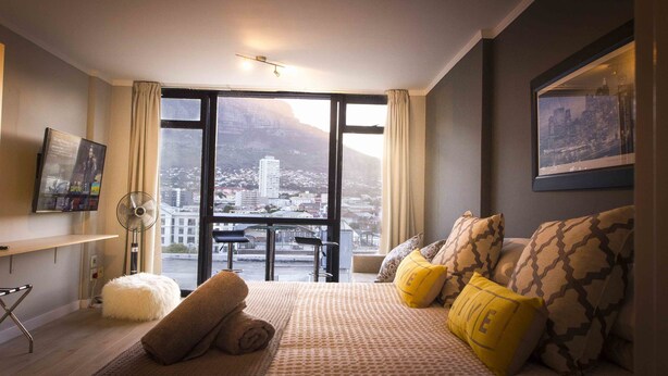 Quarto de hotel moderno com uma janela ampla, cama com roupa de cama estampada, um banquinho branco fofo e vista para a cidade.