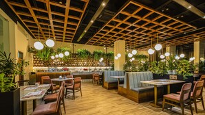 Interior de restaurante com piso de madeira, várias áreas de estar incluindo cabines e mesas, luminárias pendentes e um bar com plantas.