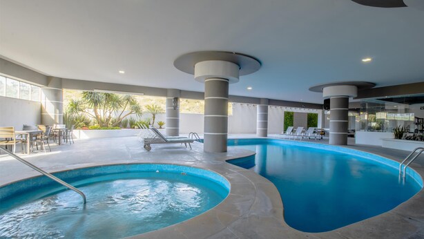 Piscina interior com duas áreas distintas, cadeiras de descanso e um espaço de estar com mesas.