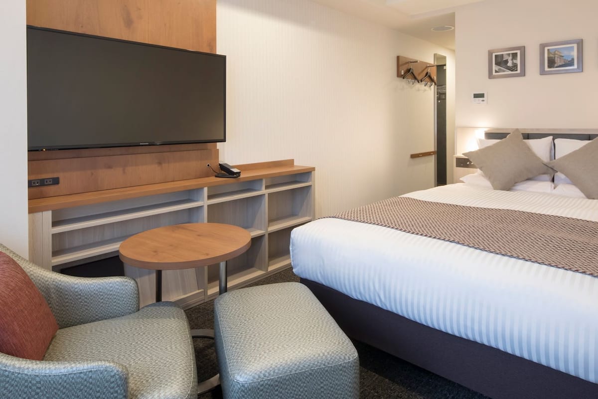 Quarto de hotel com cama, televisão na parede, mesa redonda pequena e duas poltronas.