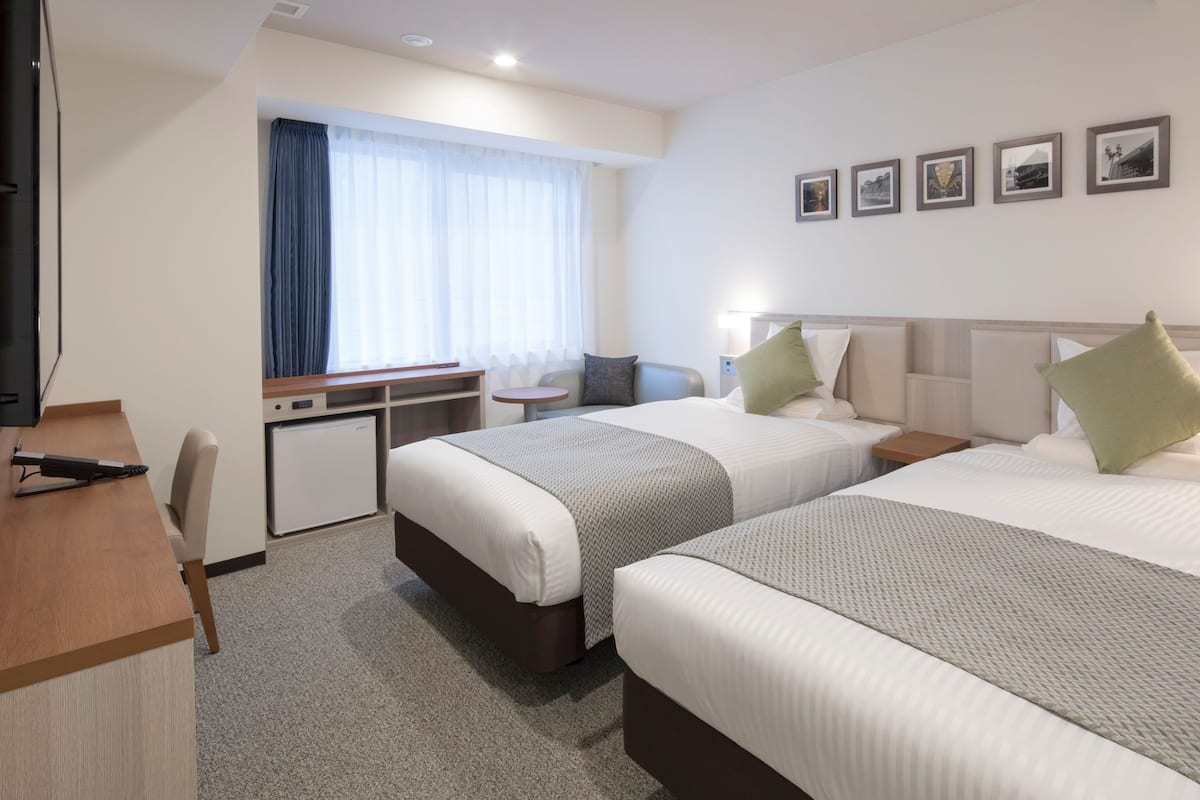 Quarto de hotel com duas camas, uma escrivaninha, uma cadeira, um pequeno frigorífico e uma televisão.