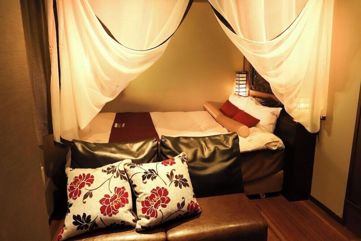 Quarto com cama, mesa de cabeceira, luminária e um banquinho com almofadas florais.