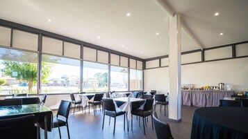 Um espaço amplo e interno com várias mesas e cadeiras, janelas grandes com vista para o exterior e um serviço de buffet de um lado.