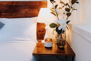 Cama bem arrumada com lençóis brancos, mesa de cabeceira de madeira com abajur branco, vaso de vidro transparente com flores brancas e uma pequena vela.