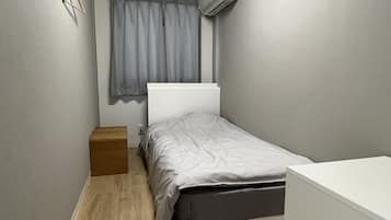 Quarto pequeno e bem organizado, com uma cama de solteiro, mesa de cabeceira e janela com cortinas.