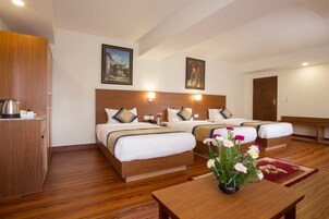 Quarto de hotel com duas camas, piso de madeira, uma escrivaninha com um vaso de flores e luminárias fixadas na parede.