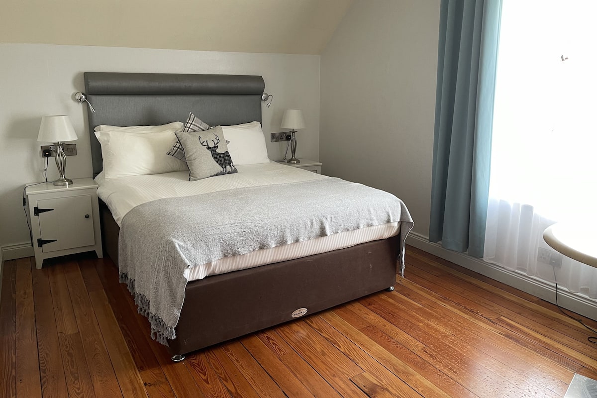 Cama bem arrumada com cabeceira cinza, roupa de cama branca e manta cinza. Duas mesas de cabeceira com luminárias e um pequeno armário. O quarto possui piso de madeira e uma janela com cortinas azuis.