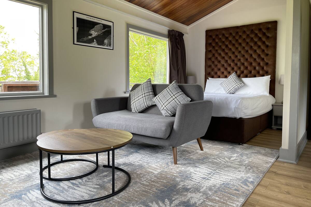 Quarto moderno com sofá cinza, mesa de centro redonda, cabeceira estofada e uma janela grande.