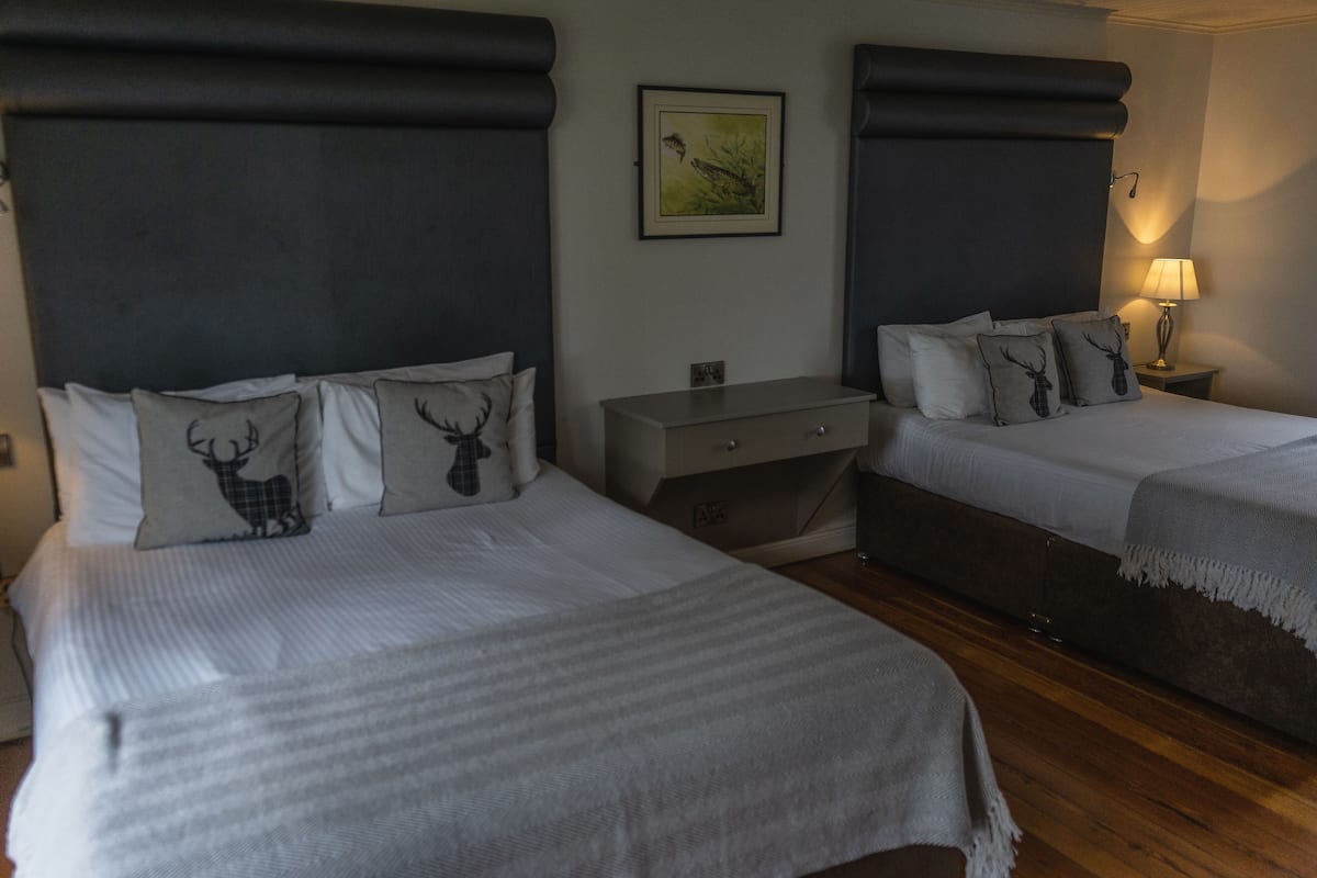 Quarto de hotel com duas camas, cada uma com cabeceira e várias almofadas, uma mesinha de cabeceira e uma luminária fixada na parede.