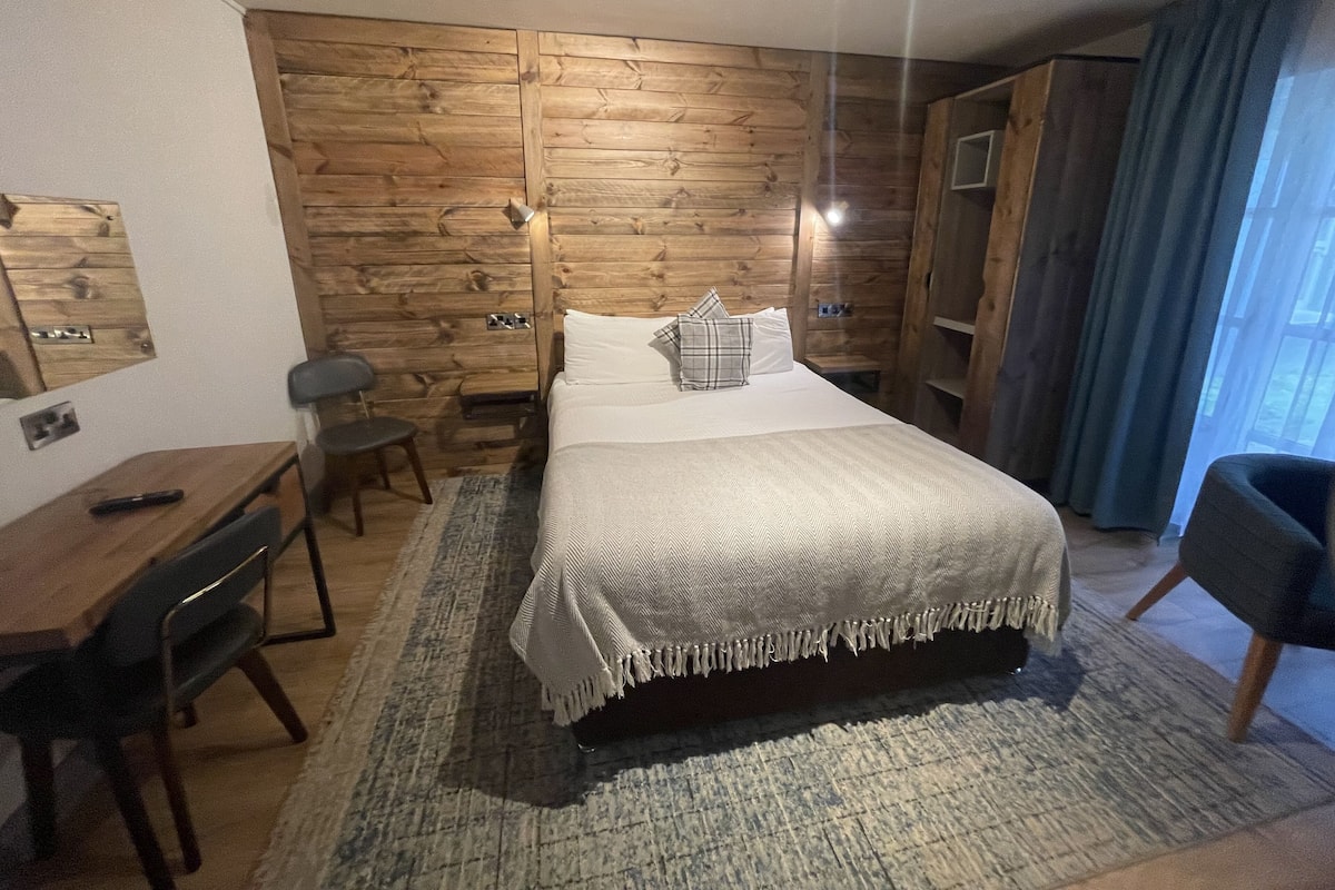 Quarto de hotel com parede de madeira, cama com colcha branca, escrivaninha com cadeira e guarda-roupa.