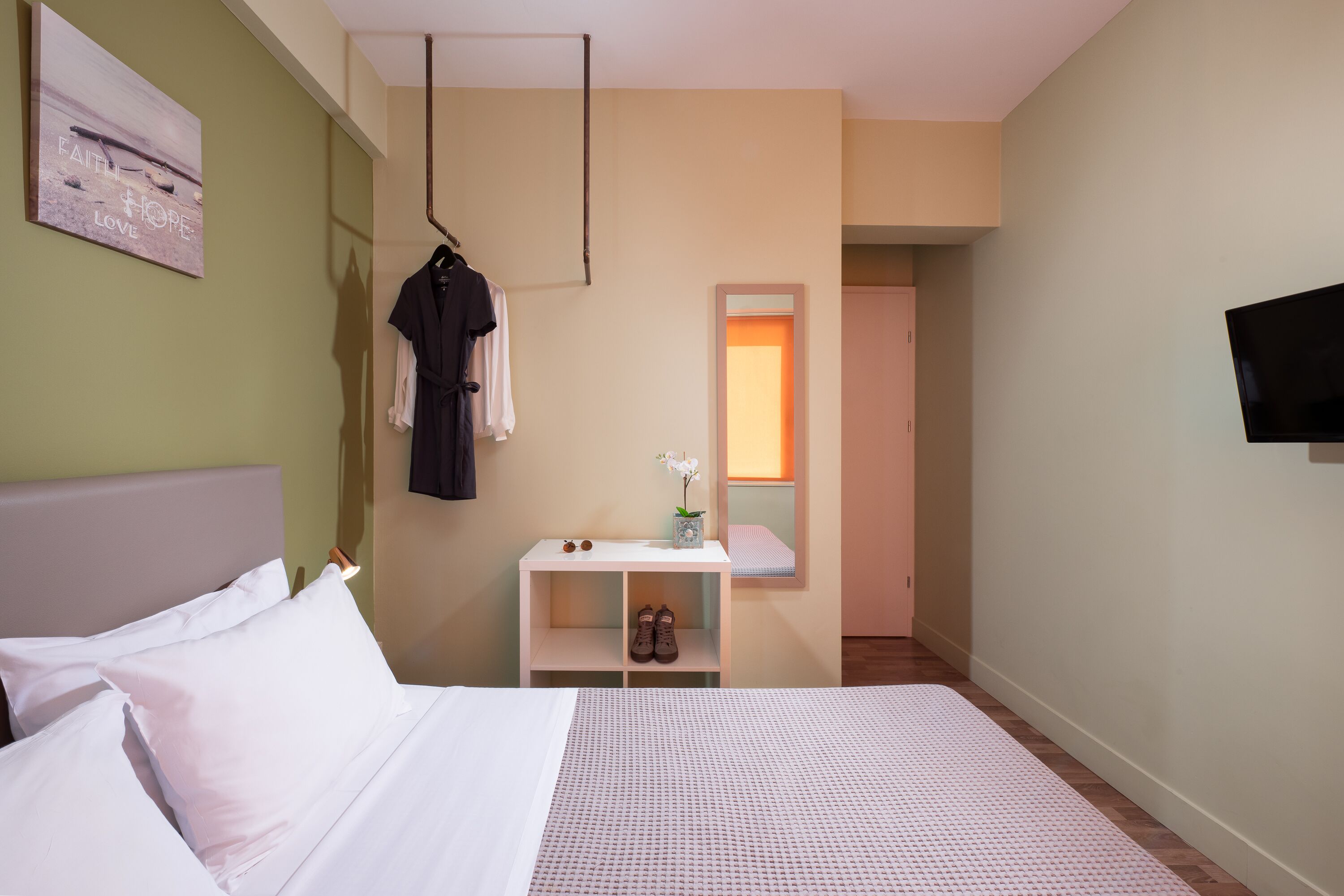 Quarto de hotel com uma cama, um roupão pendurado em um cabide, um pequeno armário branco com sapatos, um espelho e uma televisão na parede.