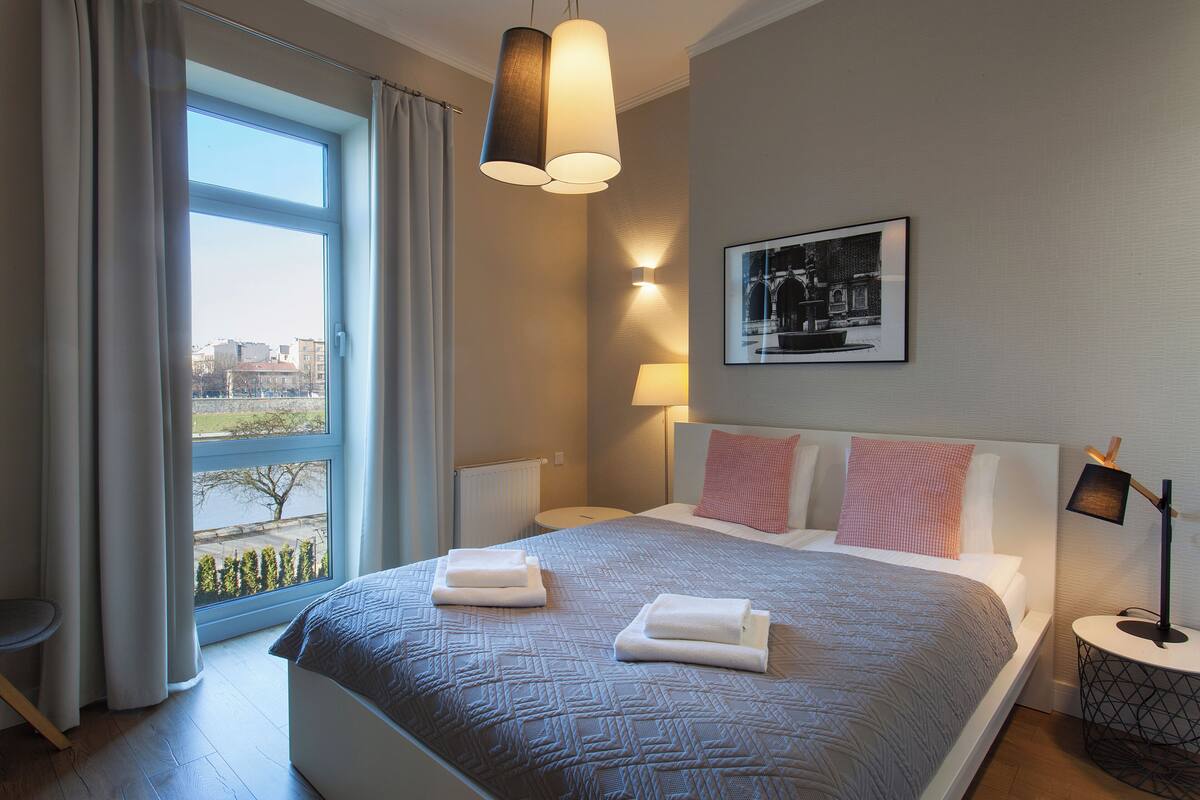 Quarto de hotel com uma cama grande, duas luminárias laterais, vista para a cidade e um quadro na parede.