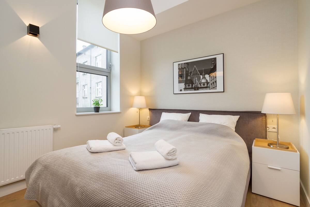 Cama bem arrumada com duas toalhas enroladas, mesa de cabeceira com abajur, quadro na parede e janela com vista para edifícios.