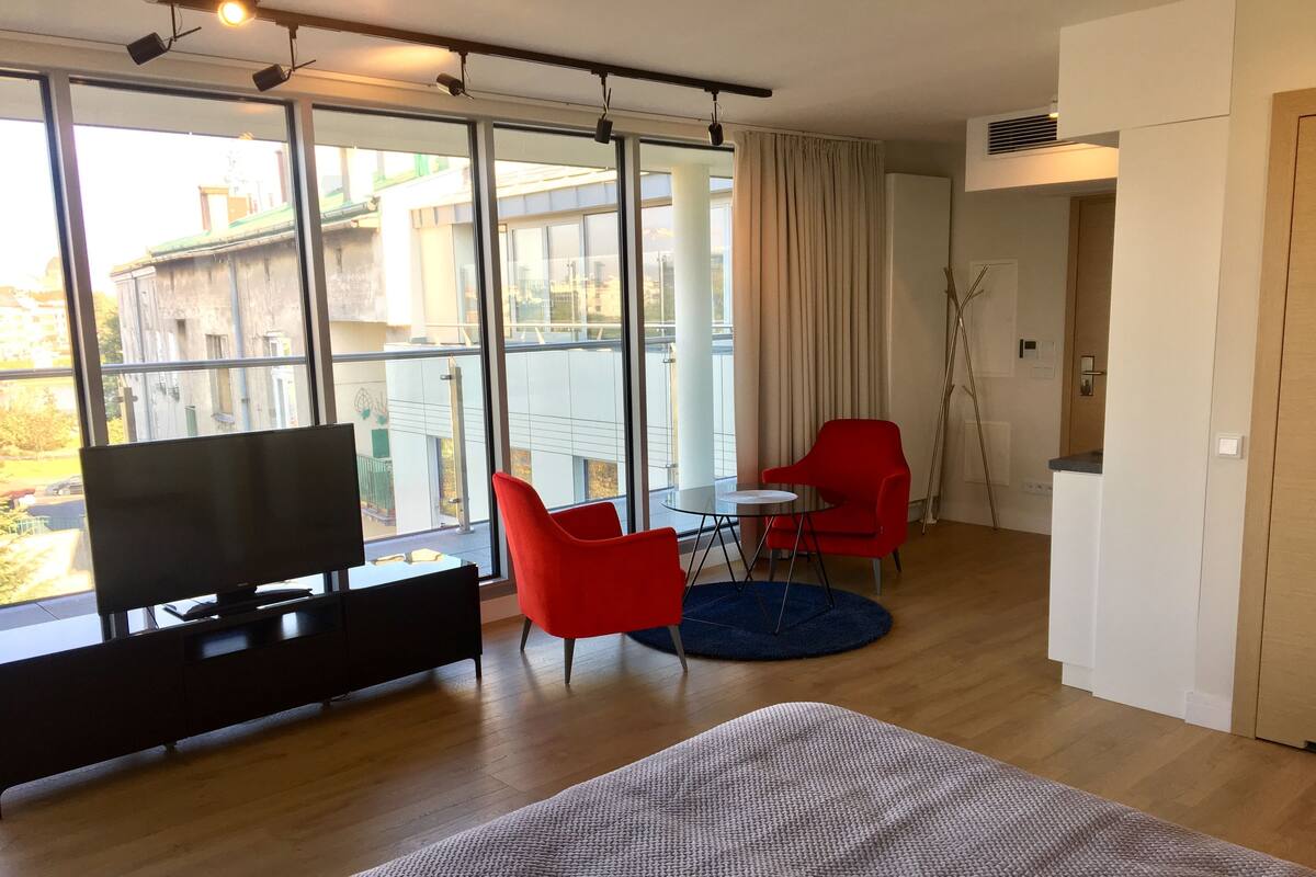 Quarto de hotel moderno com uma janela grande, televisão de tela plana, uma cadeira vermelha, uma mesa redonda pequena e uma cama.