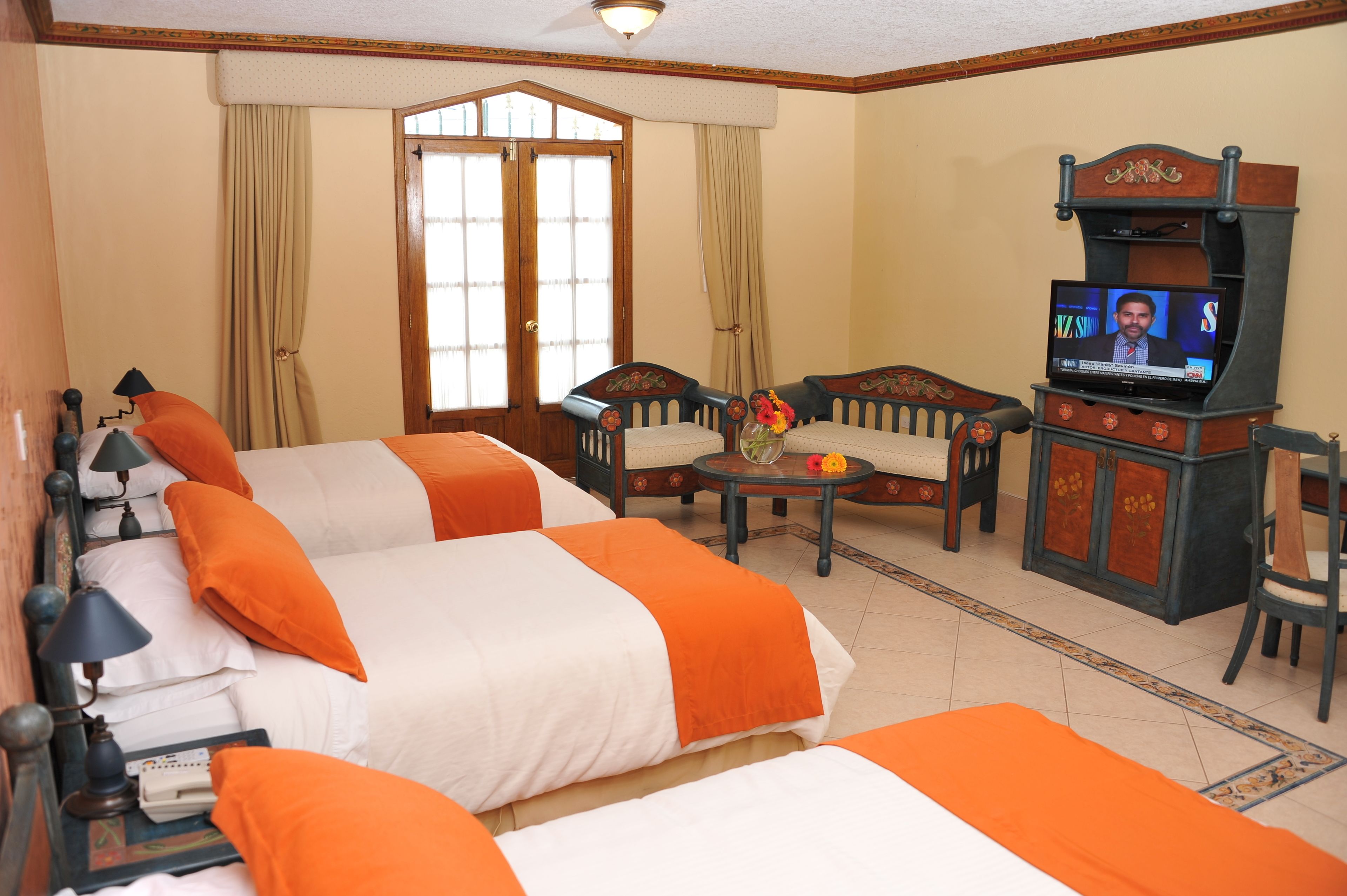 Quarto de hotel com duas camas, uma televisão, uma mesinha com um vaso e uma janela com cortinas.