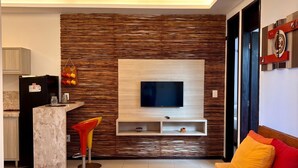Uma sala de estar moderna com televisão de tela plana fixada em uma parede de madeira, ventilador de teto, cozinha com geladeira e área de estar com sofá e almofadas coloridas.
