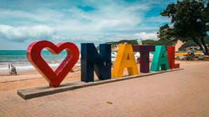 Uma praia com letras grandes e coloridas formando a palavra 'NATAL' e uma letra em formato de coração.