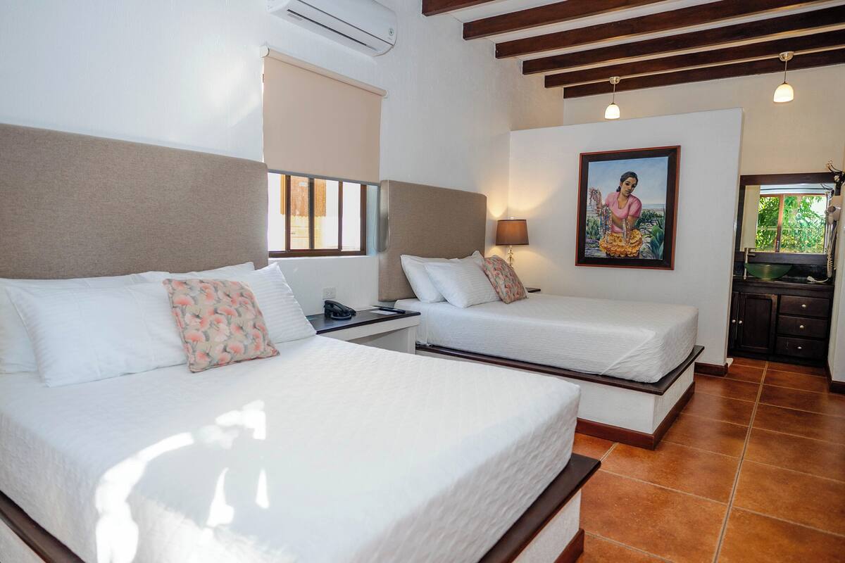 Quarto de hotel com duas camas, uma escrivaninha, uma lâmpada, um quadro e vista para o exterior.