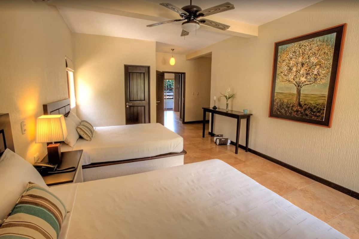 Quarto de hotel com duas camas, um ventilador de teto, um quadro de uma árvore e uma mesa com abajur.