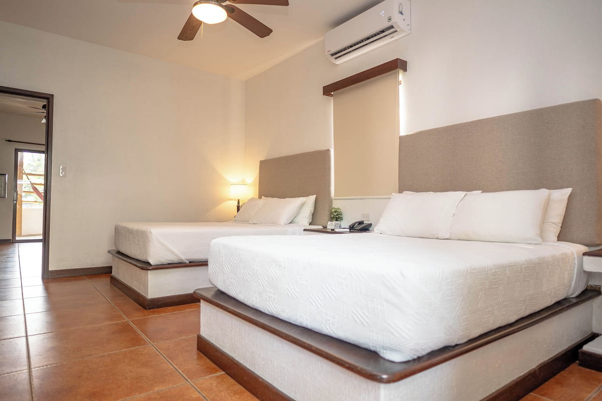 Quarto de hotel com duas camas, ventoinha no teto, ar condicionado e mesa de cabeceira com abajur.