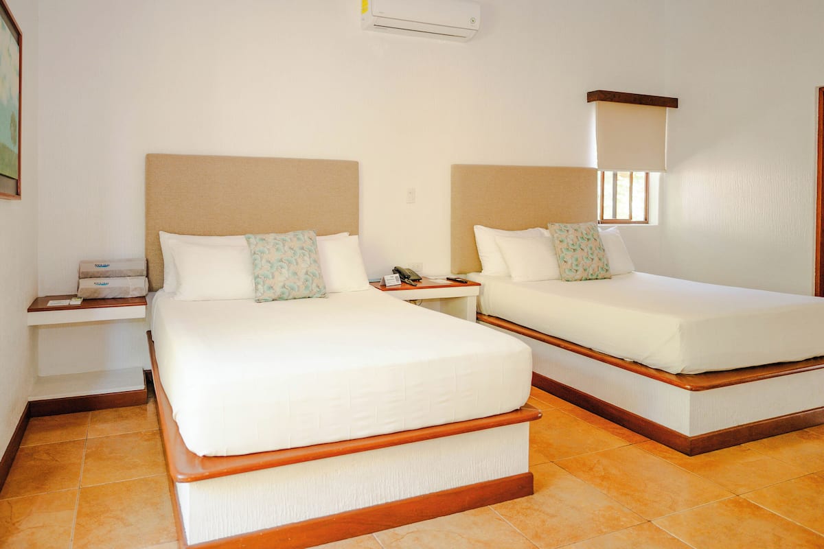 Quarto de hotel com duas camas, cada uma com cabeceira de madeira, roupa de cama branca e uma pequena mesa de cabeceira com abajur.