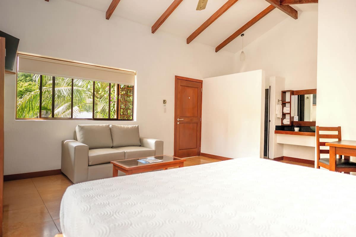 Quarto com uma cama grande, um sofá, uma mesa de centro de madeira, um guarda-roupa e uma janela com vista para o verde.
