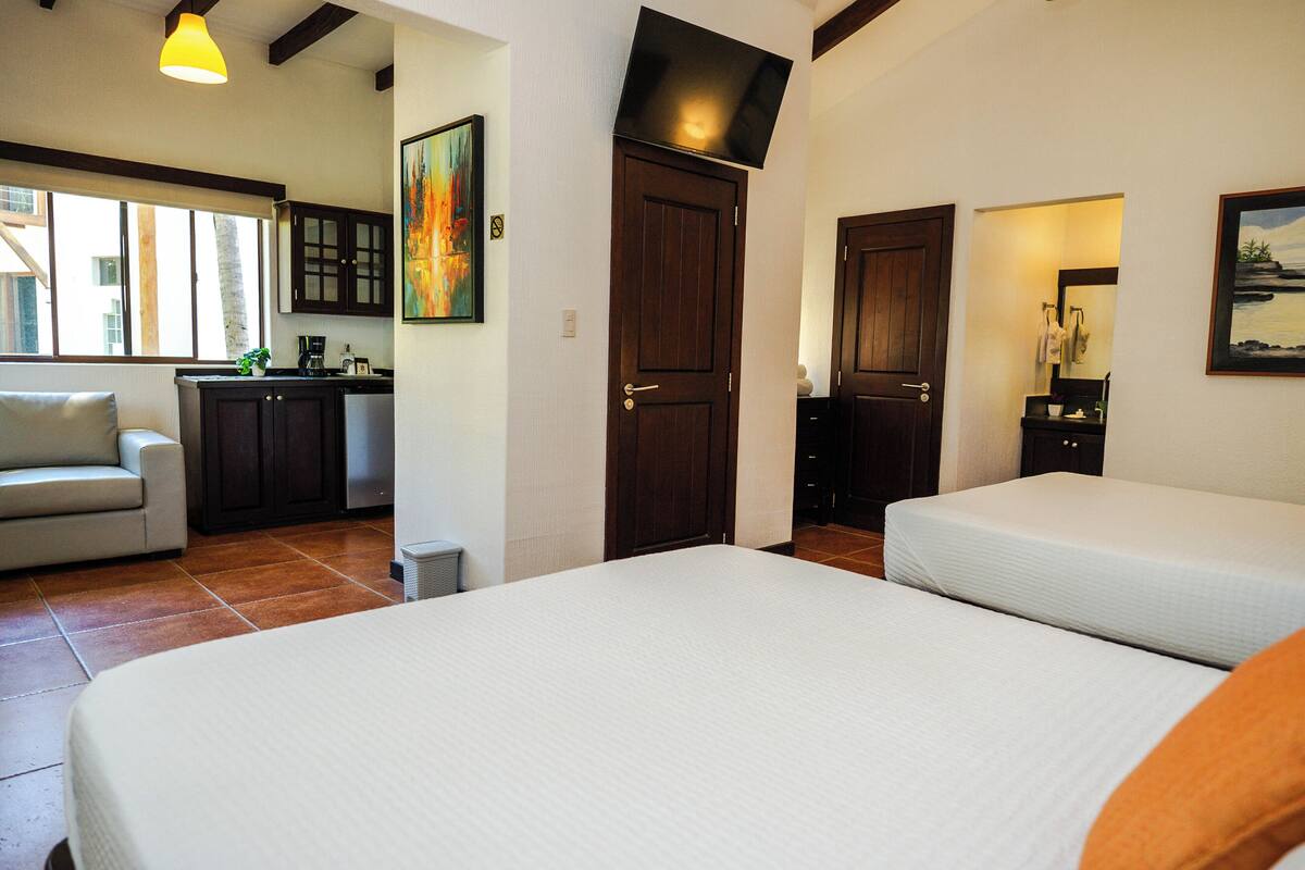Quarto de hotel com duas camas, cozinha compacta, área de refeições e uma janela grande.
