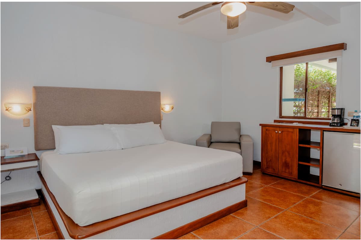Quarto de hotel com uma cama grande, uma mesa com cadeira, uma kitchenette pequena e um ventilador de teto.