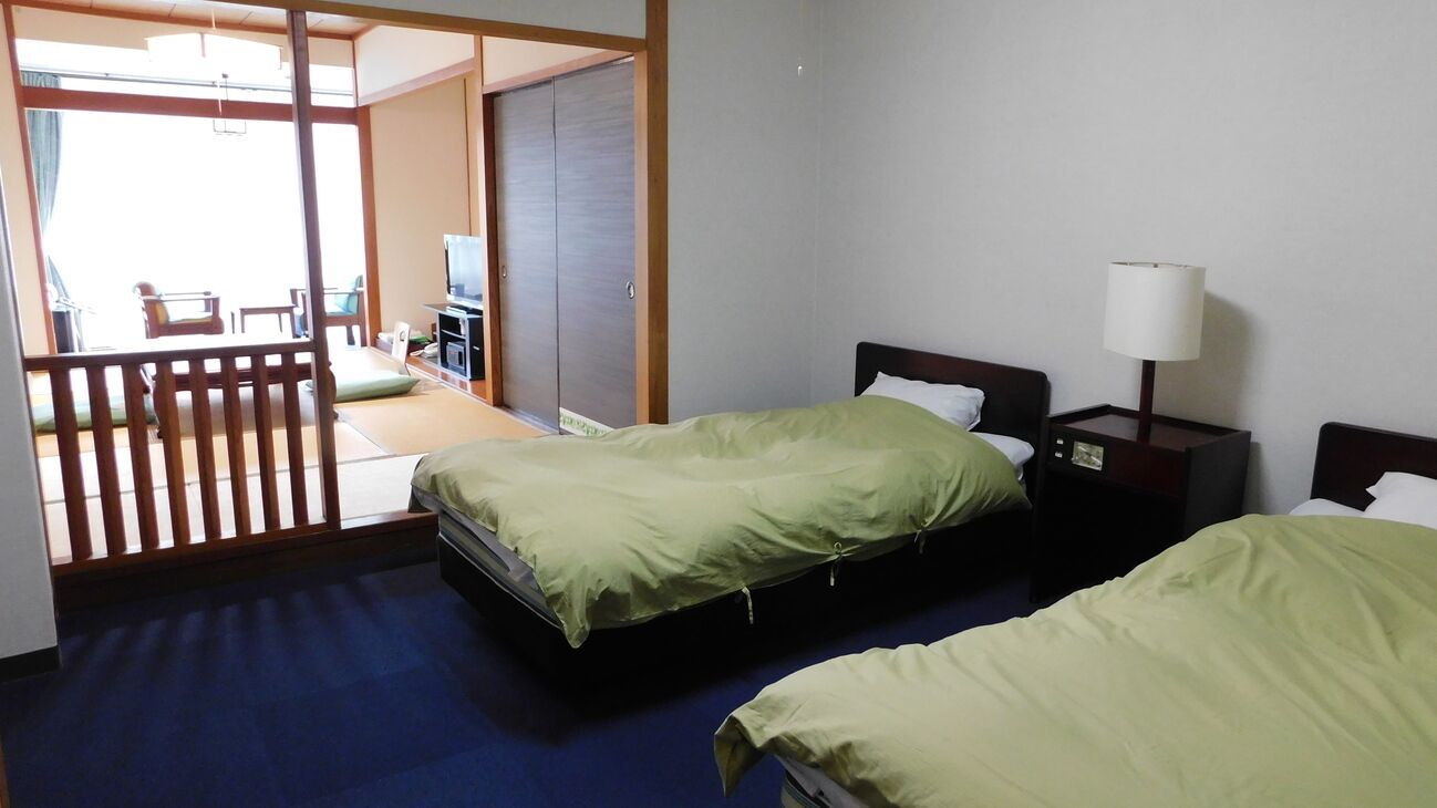 Quarto de hotel com duas camas, uma mesa, uma cadeira, uma TV e uma porta de correr que dá para uma varanda.
