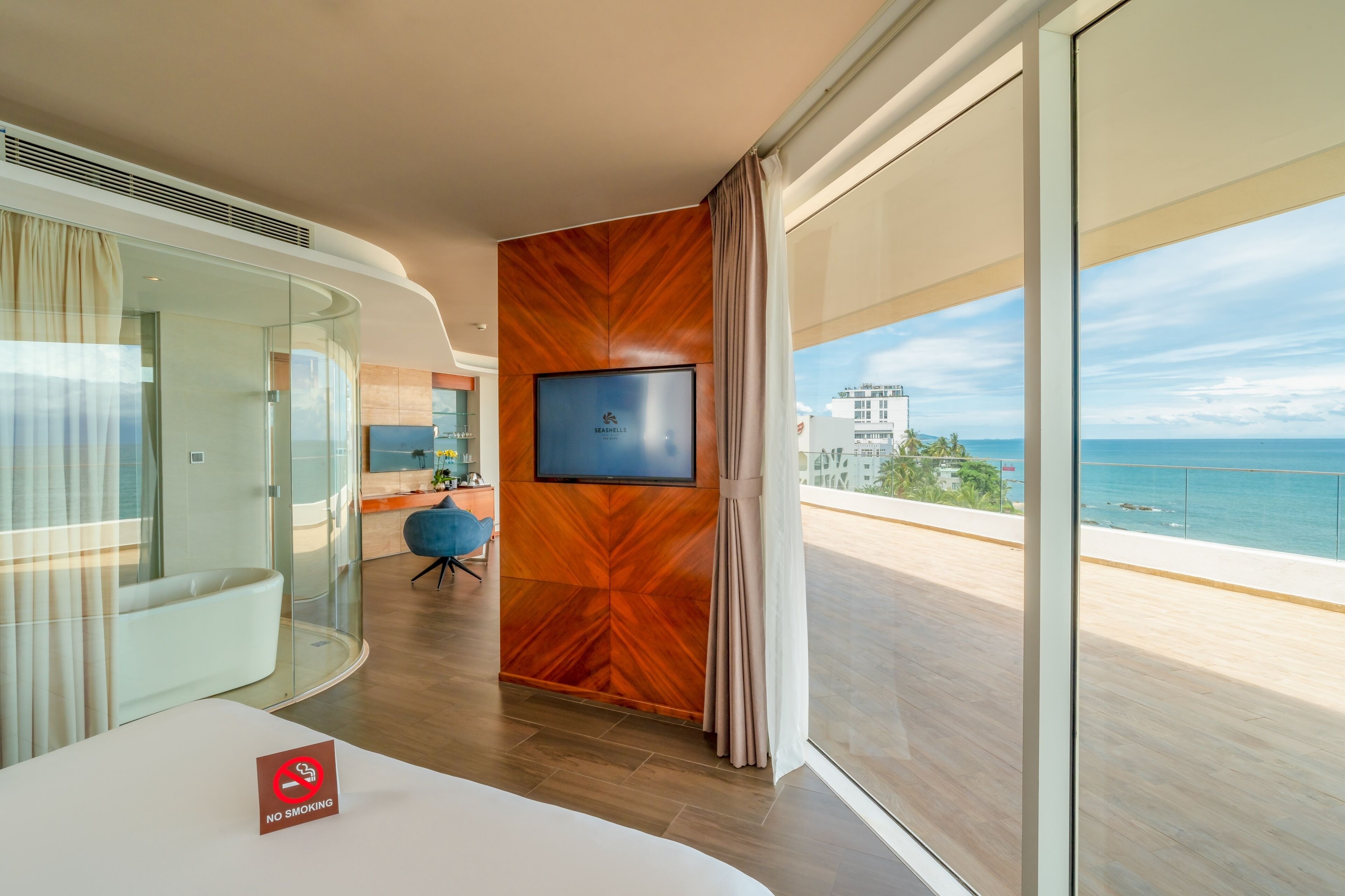 Quarto de hotel moderno com uma televisão grande, box de chuveiro de vidro, banheira branca, uma cadeira azul e uma varanda com vista para o mar.