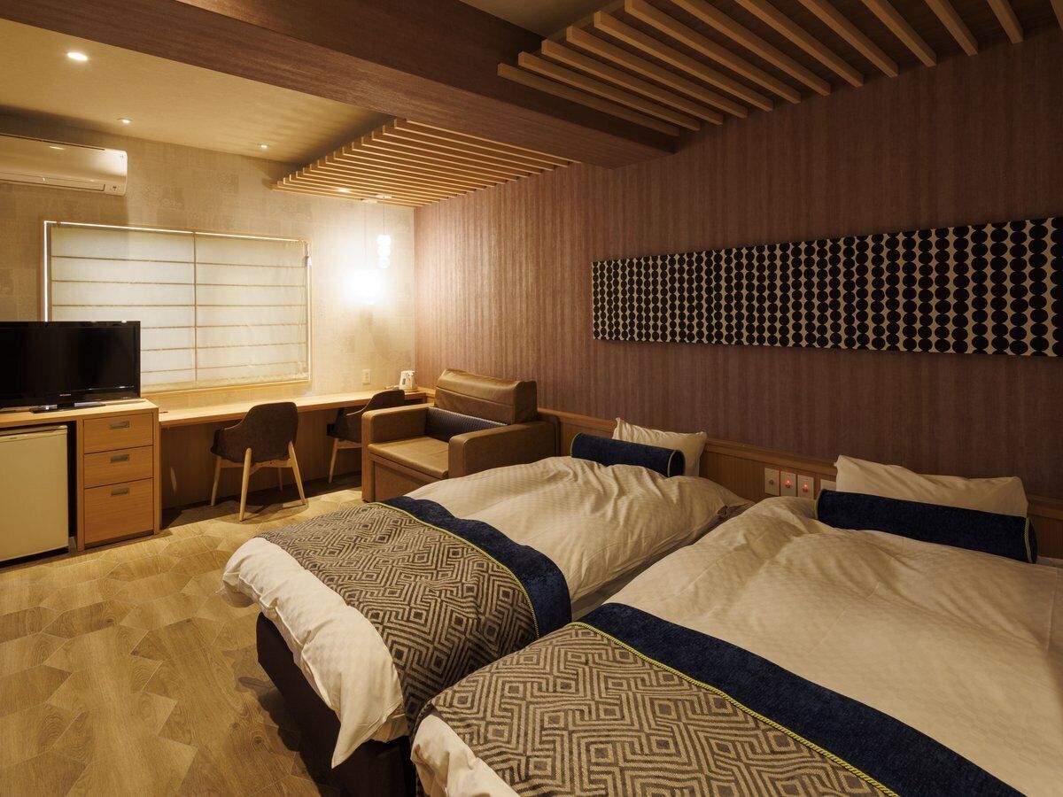 Quarto de hotel com duas camas, uma escrivaninha, uma cadeira, uma televisão e um candeeiro fixo na parede.