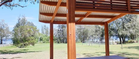 Um pavilhão de madeira com telhado de metal corrugado, um banco e um campo desportivo cercado ao fundo.