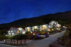 Uma casa de vários andares com uma escadaria que leva à entrada, cercada por árvores e uma paisagem montanhosa.