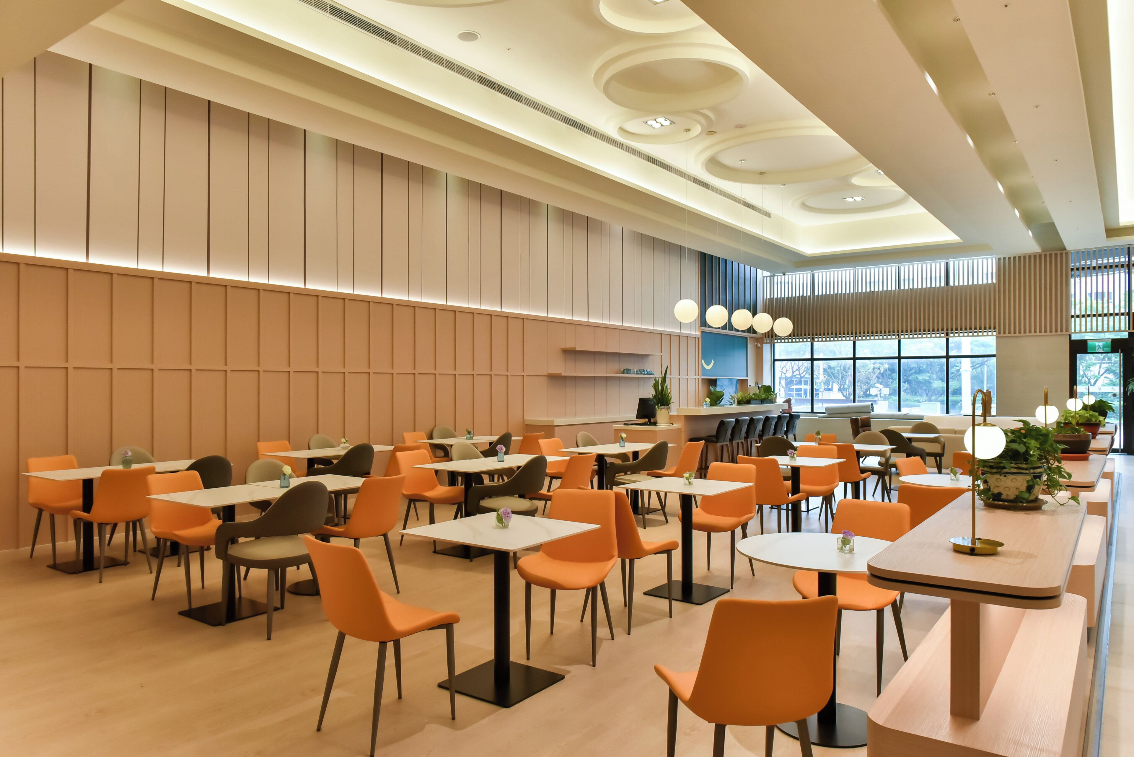 Uma sala de jantar moderna com várias mesas e cadeiras, teto alto com padrões circulares e janelas amplas que oferecem iluminação natural.
