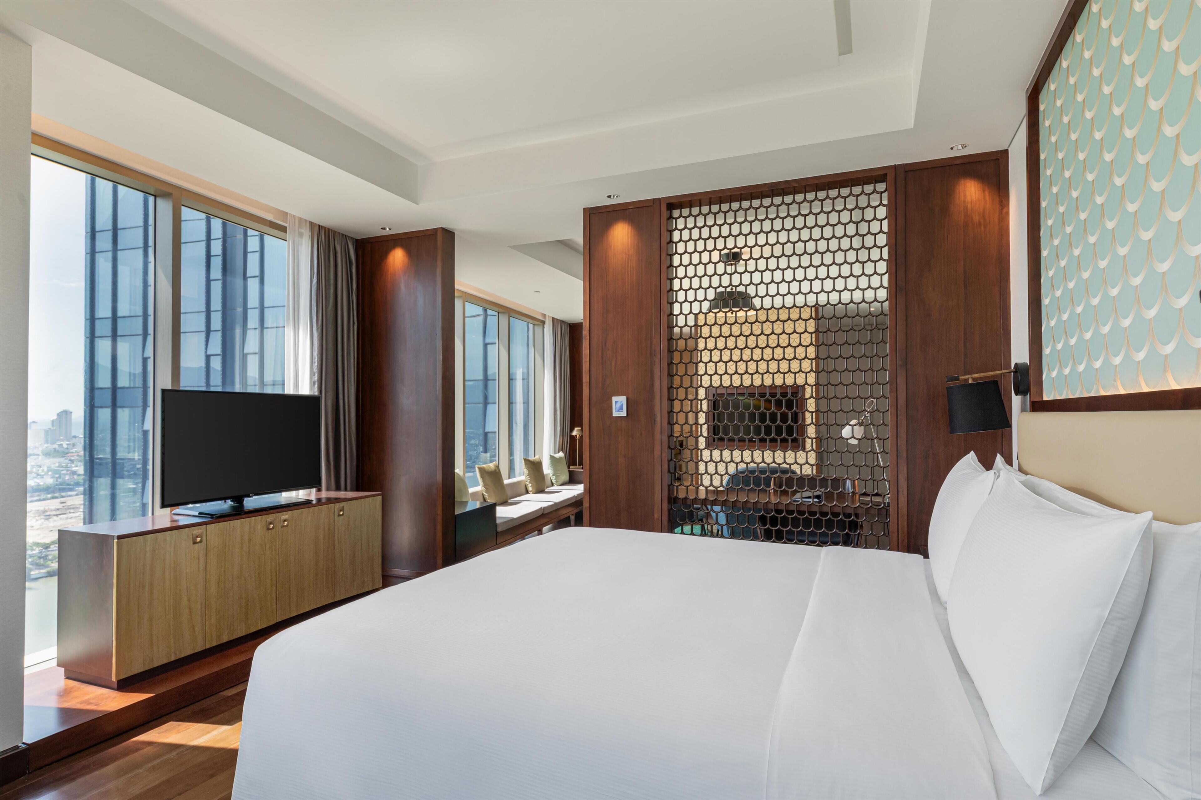Quarto de hotel moderno com uma cama grande, uma TV em um armário de madeira e vista da cidade através de janelas amplas.