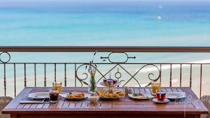 Mesa de madeira posta com pratos, copos e um vaso de flores numa varanda com vista para o oceano.