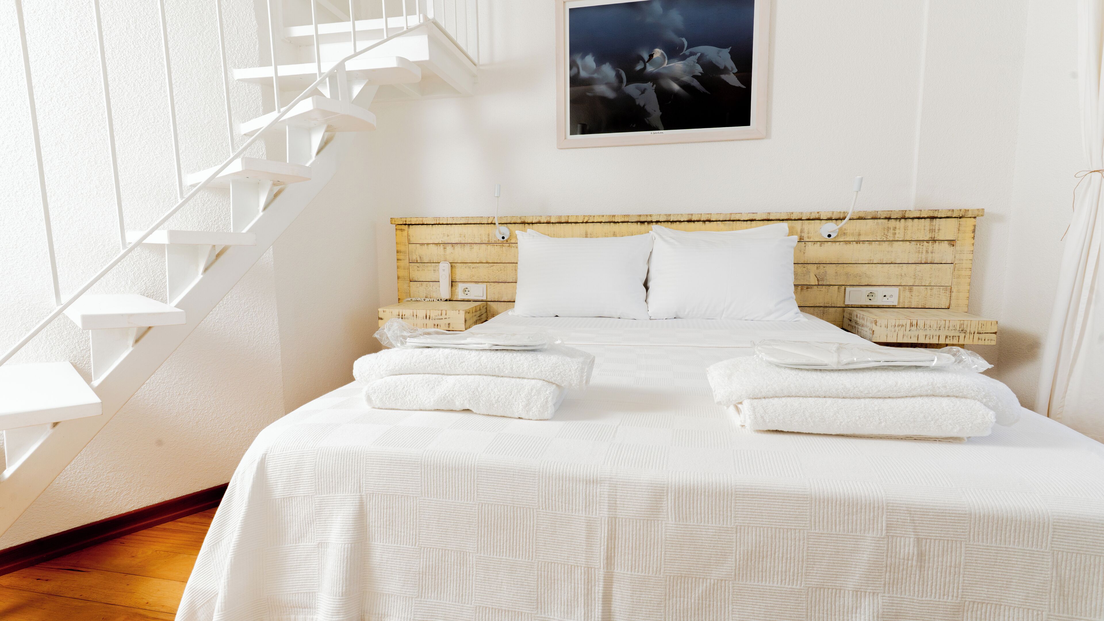 Cama bem arrumada com lençóis e toalhas brancas, cabeceira de madeira e uma escada branca à esquerda.
